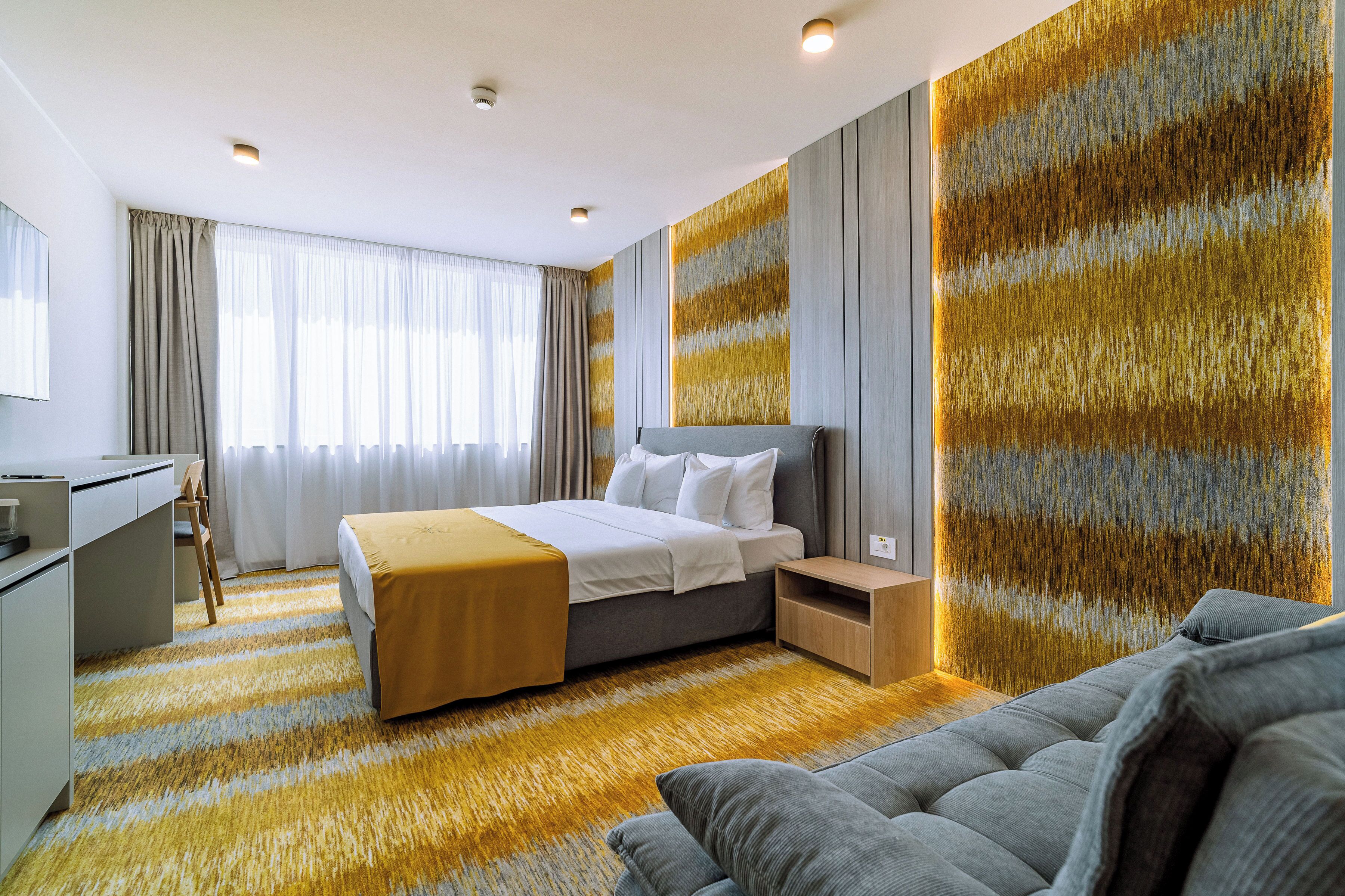 Quarto de hotel moderno com cama, sofá, escrivaninha e uma janela grande com cortinas.