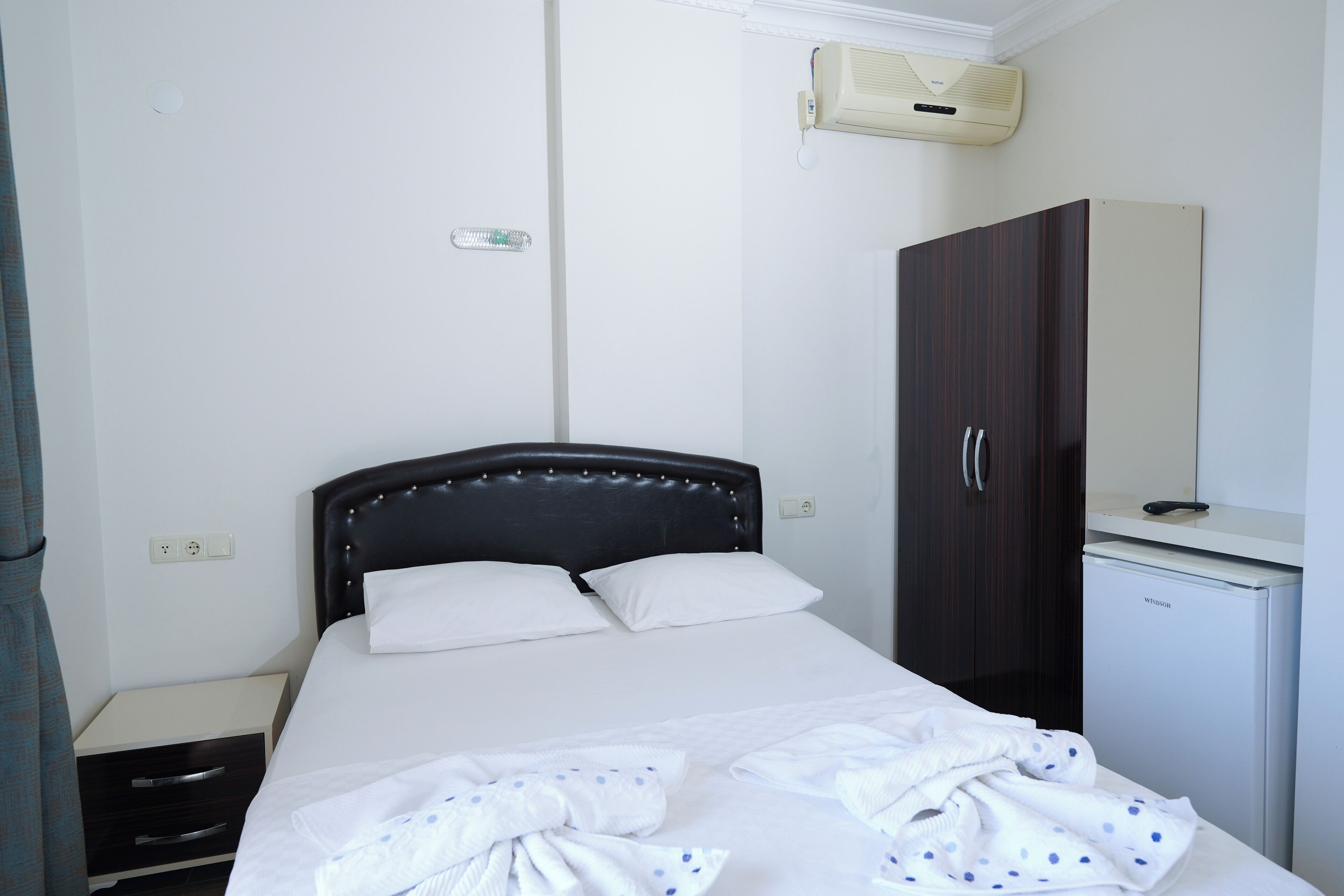 Cama bem arrumada com roupa de cama branca e manta com bolinhas, guarda-roupa de madeira escura, mesa de cabeceira pequena e branca e ar-condicionado fixado na parede.