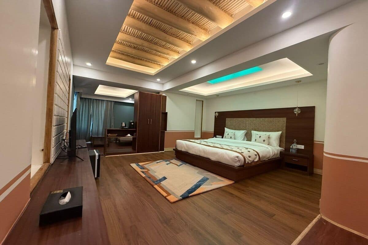Quarto de hotel moderno com uma cama grande, piso de madeira, teto com iluminação embutida e um banheiro visível ao fundo.