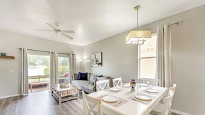 Uma sala de jantar com uma mesa branca posta para quatro pessoas, um ventilador de teto e vista para um jardim através de portas de vidro.