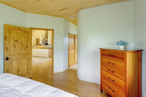 Quarto com cômoda de madeira, uma cama e um corredor que dá para uma área de cozinha.