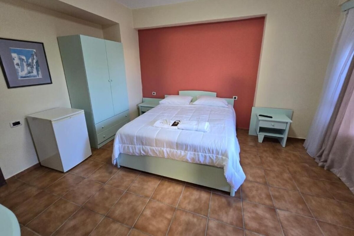 Quarto com cama, guarda-roupa, pequeno frigorífico e mesa de cabeceira.