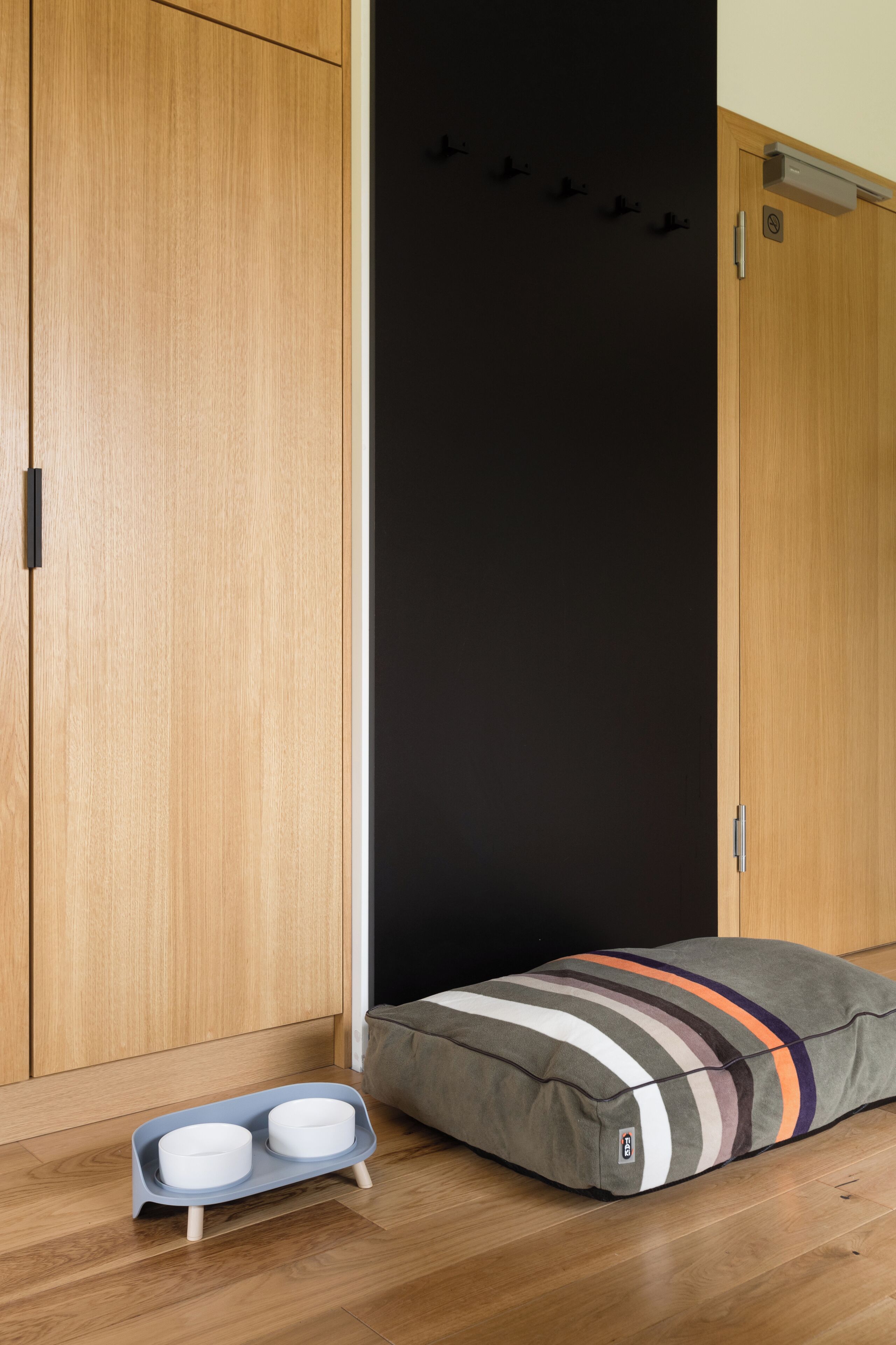 Quarto com guarda-roupa de madeira, porta preta, cama para animais de estimação cinza com listras e um comedouro branco em suporte.