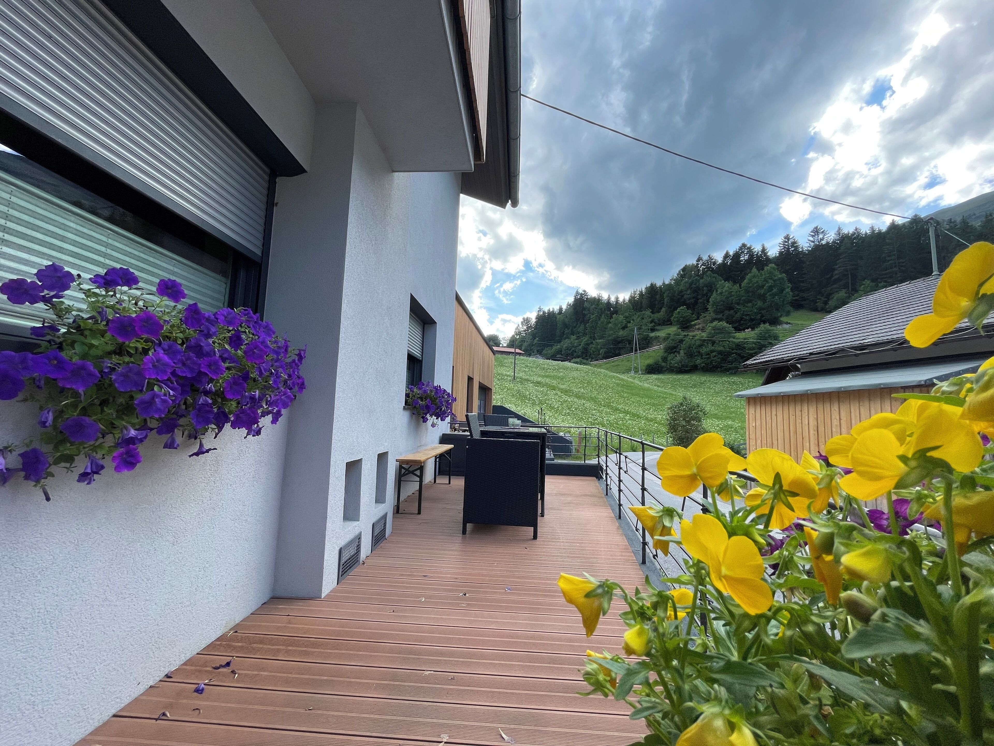Um deck de madeira com um banco e uma mesa, rodeado por um edifício branco com flores roxas penduradas e vista para uma encosta verde.