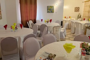 Uma área de refeições de hotel com mesas redondas cobertas por toalhas brancas, cadeiras brancas e centros de mesa.