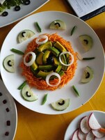 Um prato branco com cenoura ralada, fatias de abacate, fatias de pepino e vagem sobre uma mesa amarela.