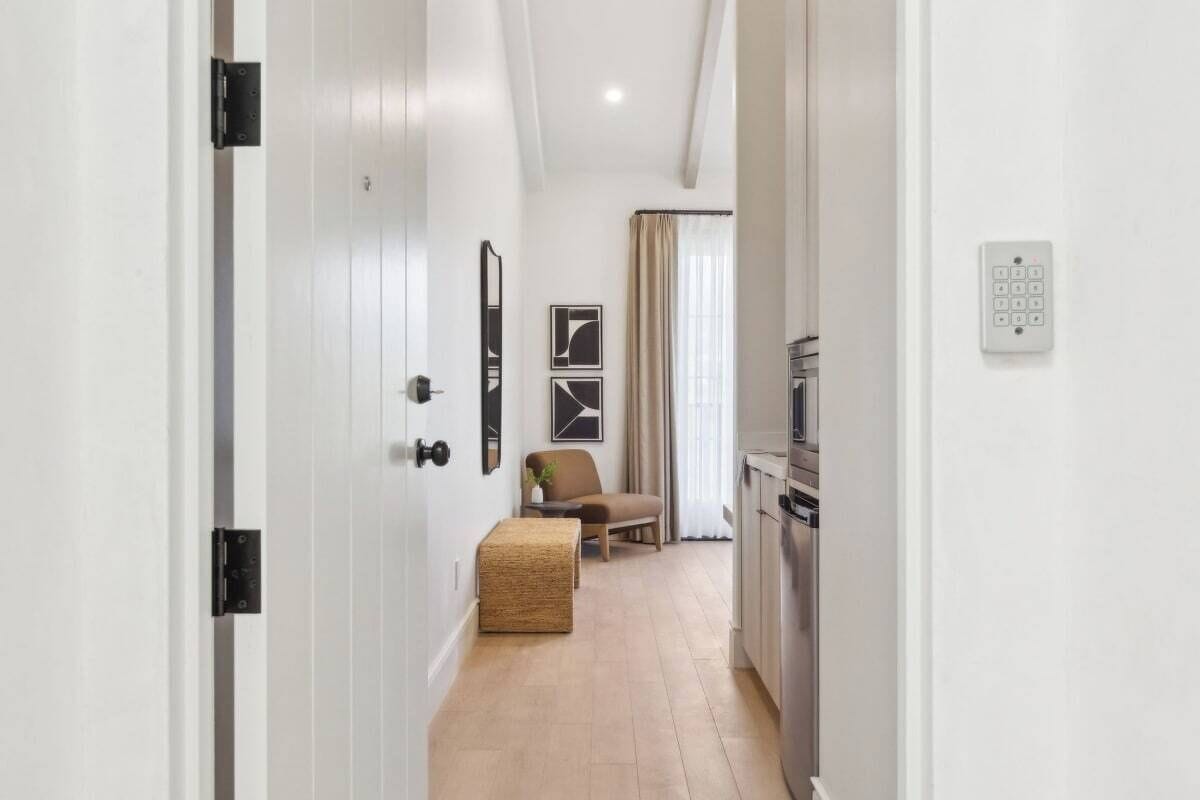 Um corredor estreito com uma porta à esquerda, um banco e uma cadeira no centro. Há uma área de cozinha à direita, com armários e um micro-ondas.