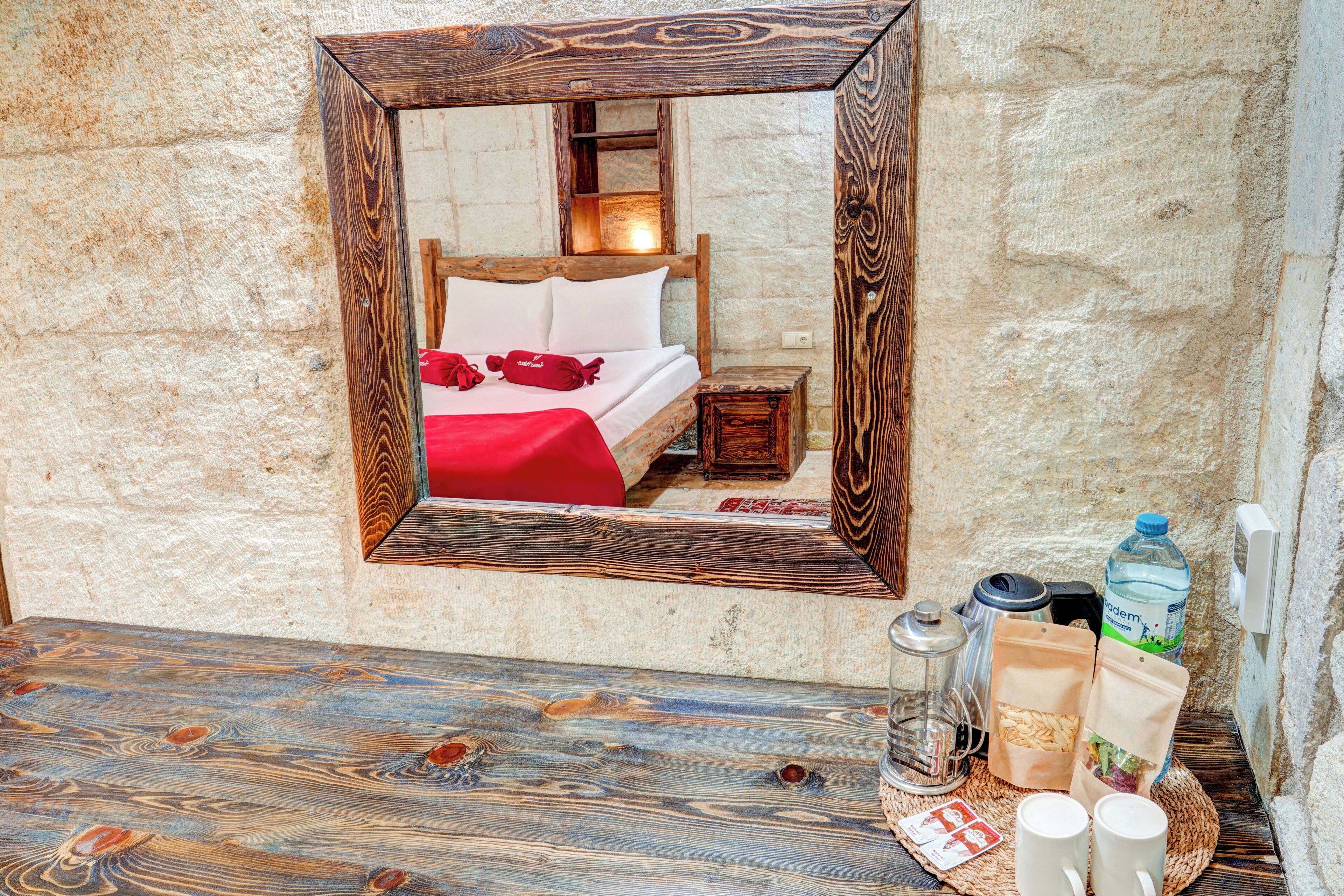 Quarto com cama de madeira, mesa de cabeceira, espelho e pequena mesa com garrafa de água, pacote de lanches e duas canecas.