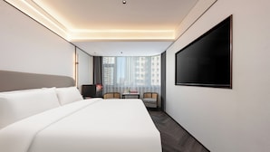 Quarto de hotel com uma cama grande, uma TV de tela plana, uma escrivaninha com cadeira e vista para edifícios altos pela janela.