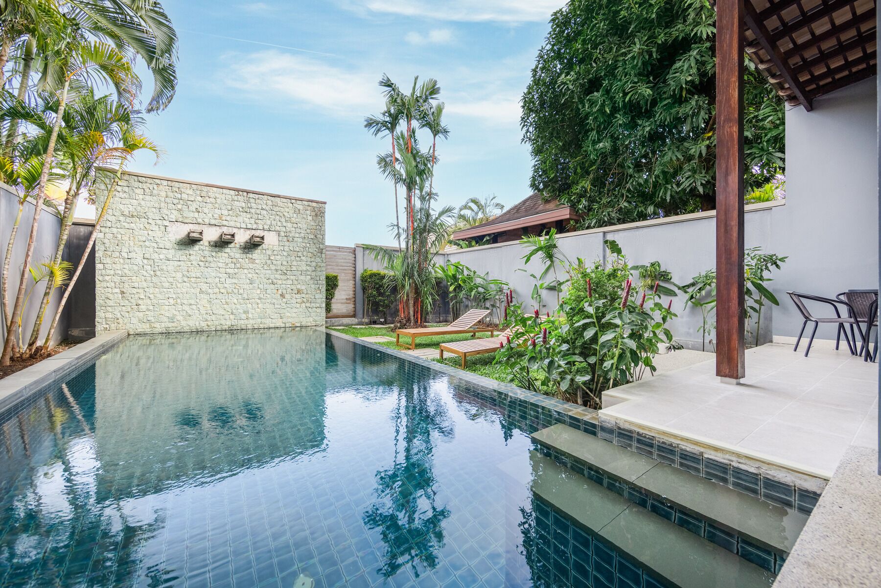 Uma piscina com muro de pedra, espreguiçadeiras de madeira e plantas tropicais.