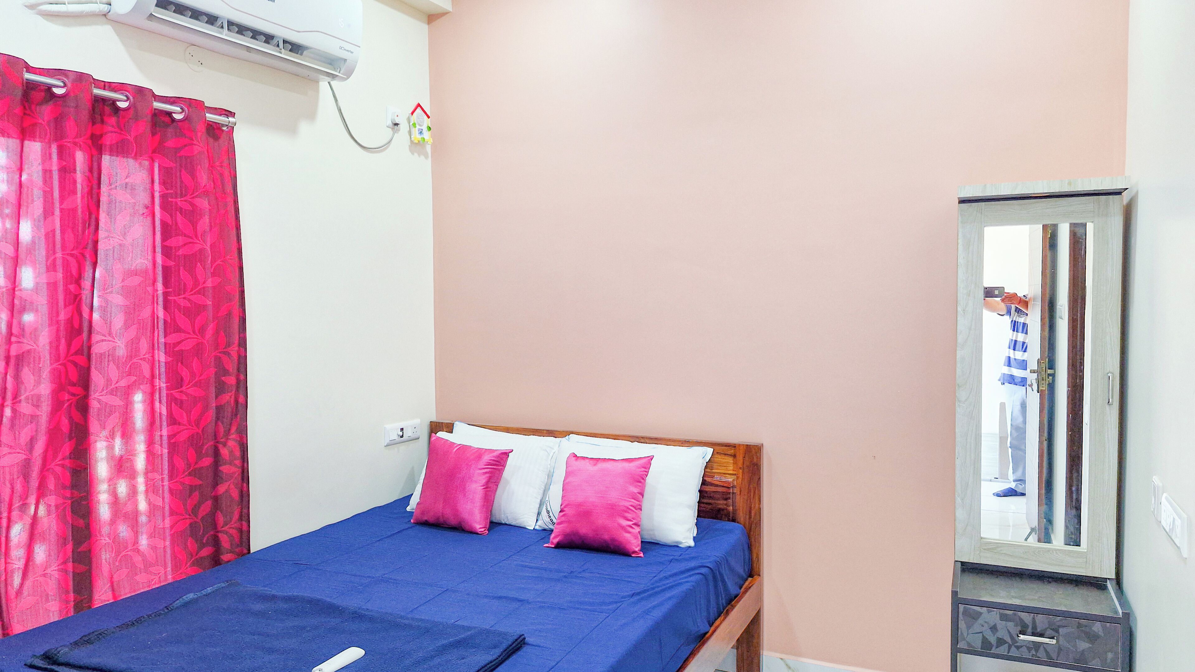 Quarto com cama, almofadas rosa e branca, colcha azul, cabeceira de madeira, espelho e ar-condicionado fixado na parede.