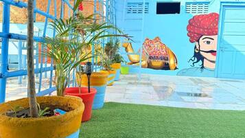 Área externa colorida com plantas em vasos, um mural e um edifício azul com uma placa que diz 'KITCHEN Staff Office'.