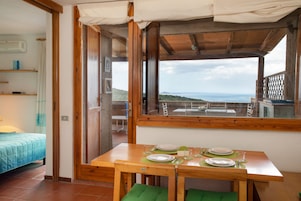 Quarto com mesa de madeira posta para refeição, cama com colcha azul e vista para o mar através de uma grande janela.