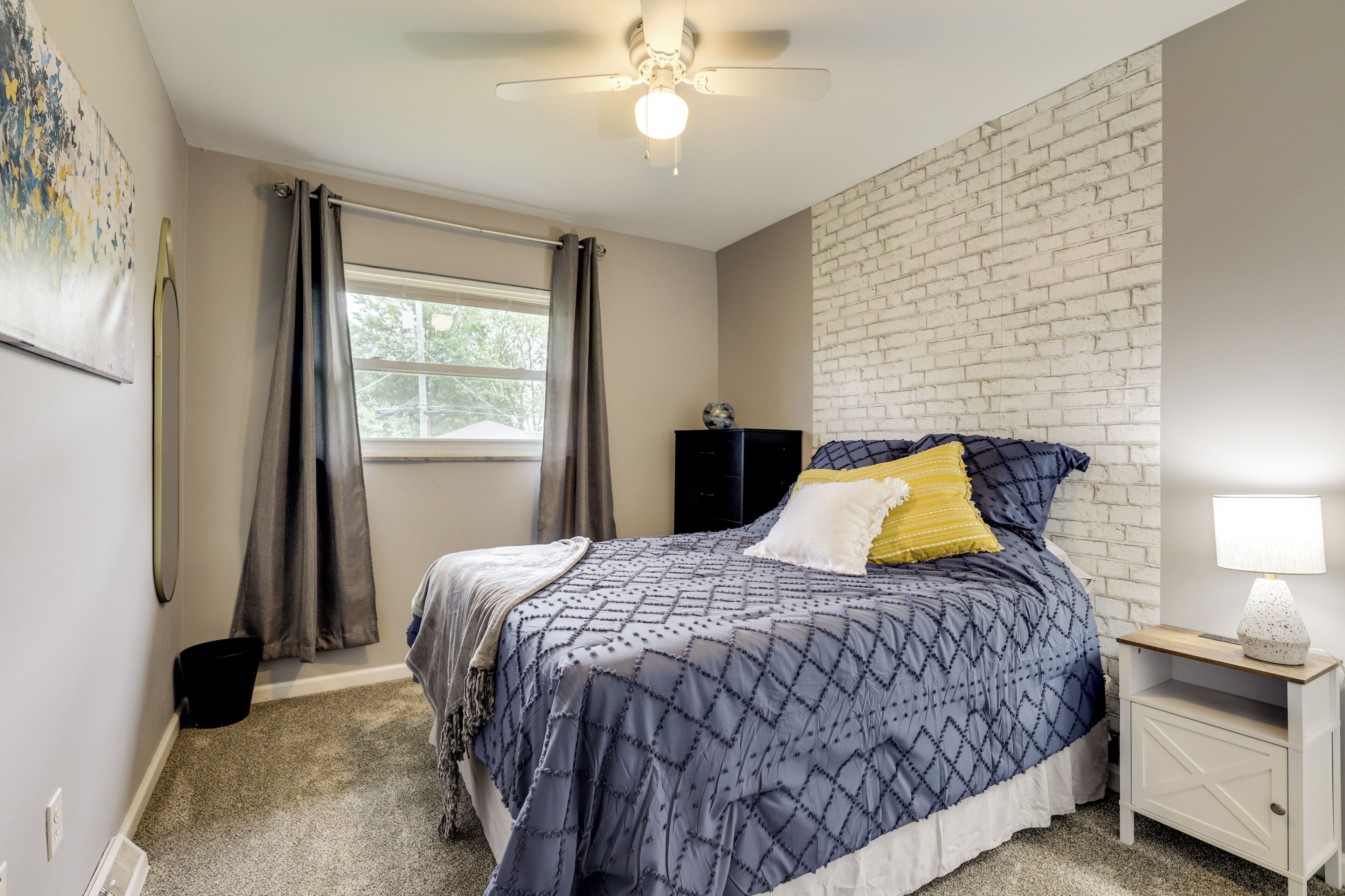 Quarto com cama, mesa de cabeceira, ventilador de teto, janela com cortinas e parede de tijolos.