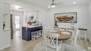 Cozinha moderna com armários brancos, uma ilha central e uma área de jantar com cadeiras brancas e mesa de madeira.