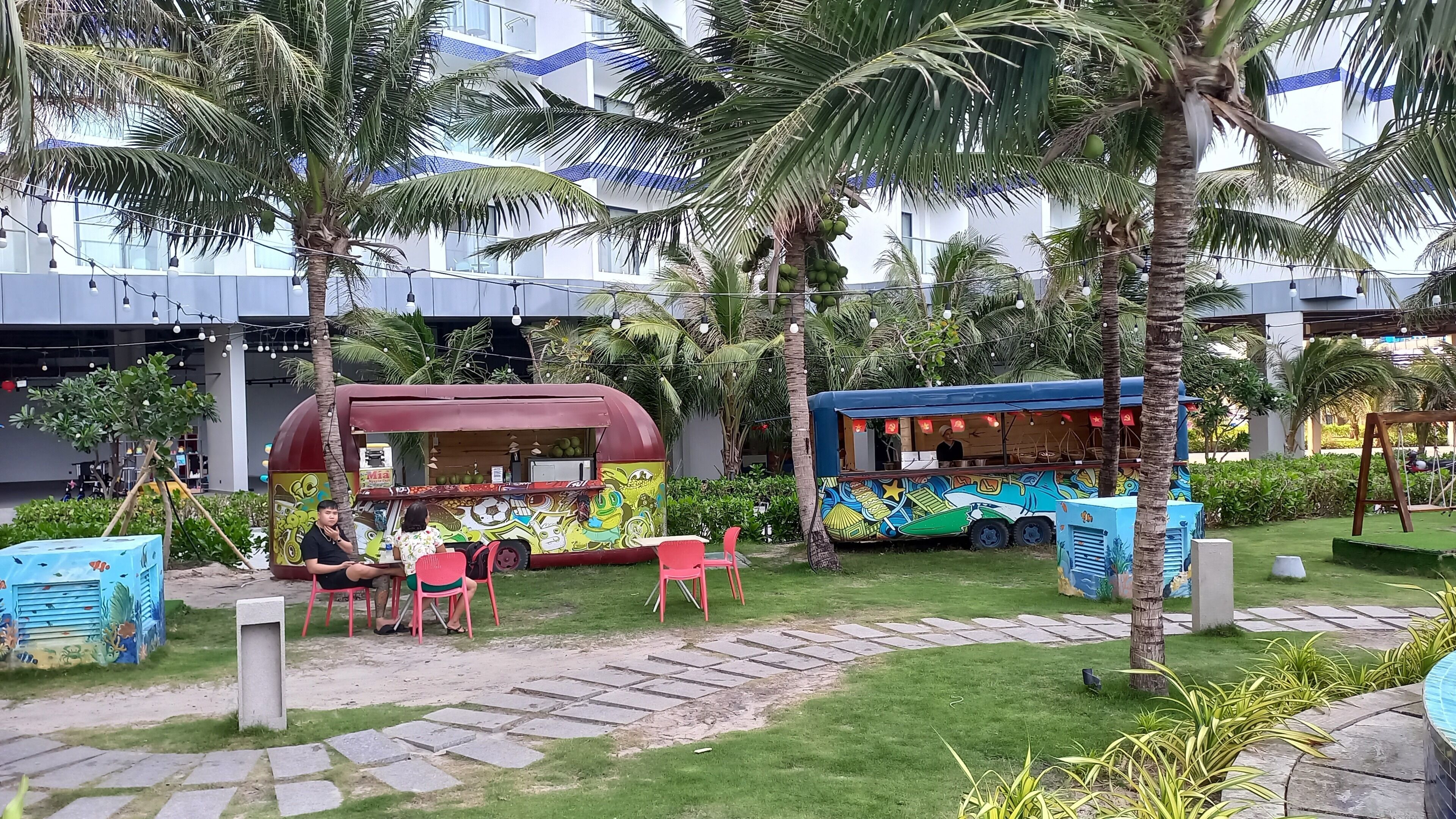 Um pátio com palmeiras, um food truck colorido e um ônibus com um mural.