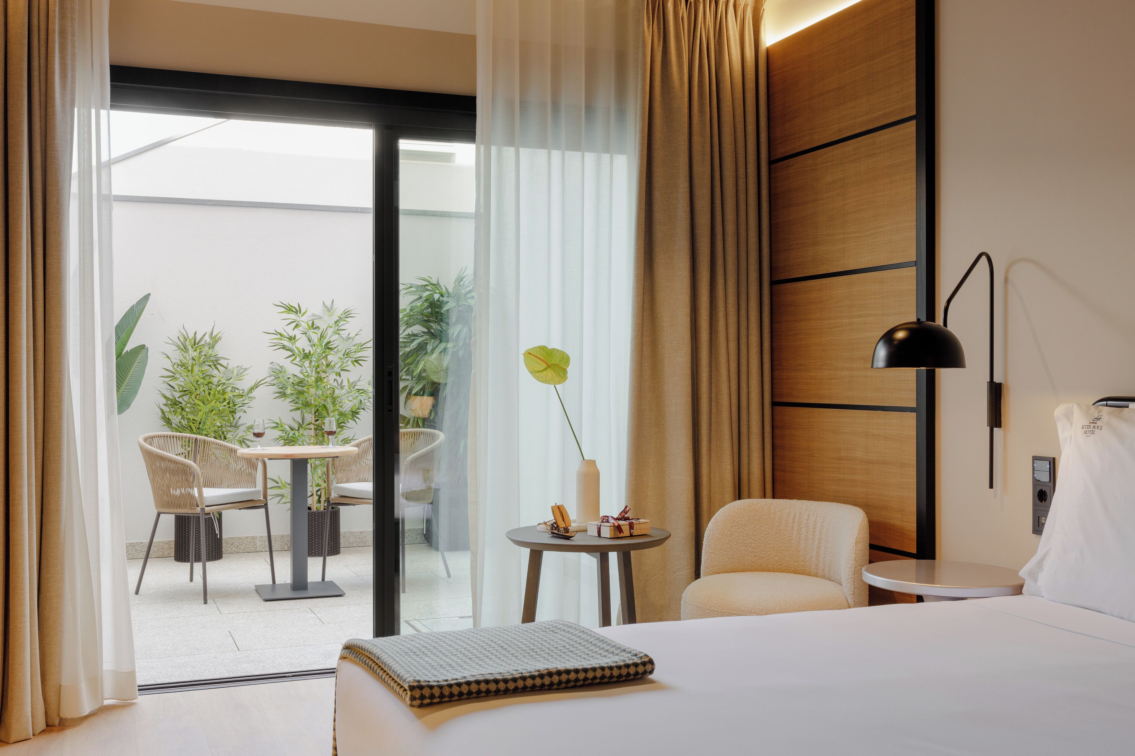 Quarto de hotel com cama, uma pequena mesa com um vaso e planta, uma cadeira, e vista para a varanda externa com plantas e uma mesa.