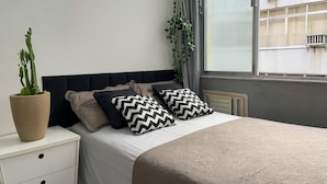 Uma cama bem arrumada com almofadas estampadas, uma planta em vaso e uma mesa de cabeceira com gavetas.