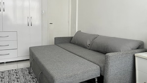 Quarto compacto com um sofá-cama cinza, um guarda-roupa branco e uma pequena mesa de cabeceira.