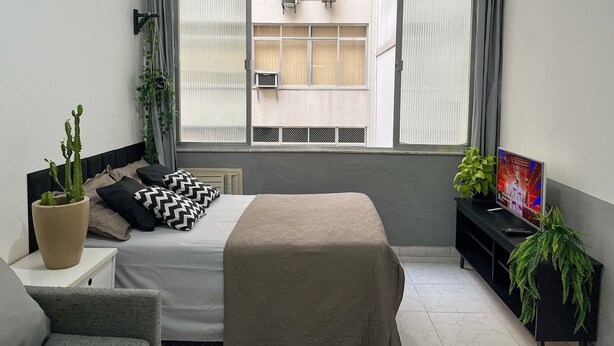 Quarto com cama, janela com cortinas, televisão e um vaso com planta.