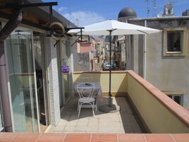 Uma pequena varanda com mesa e cadeira, um guarda-sol branco e vista para a cidade.