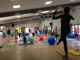 Um grupo de pessoas se exercitando com bolas de ginástica em um espaço amplo, com janelas grandes e teto alto.
