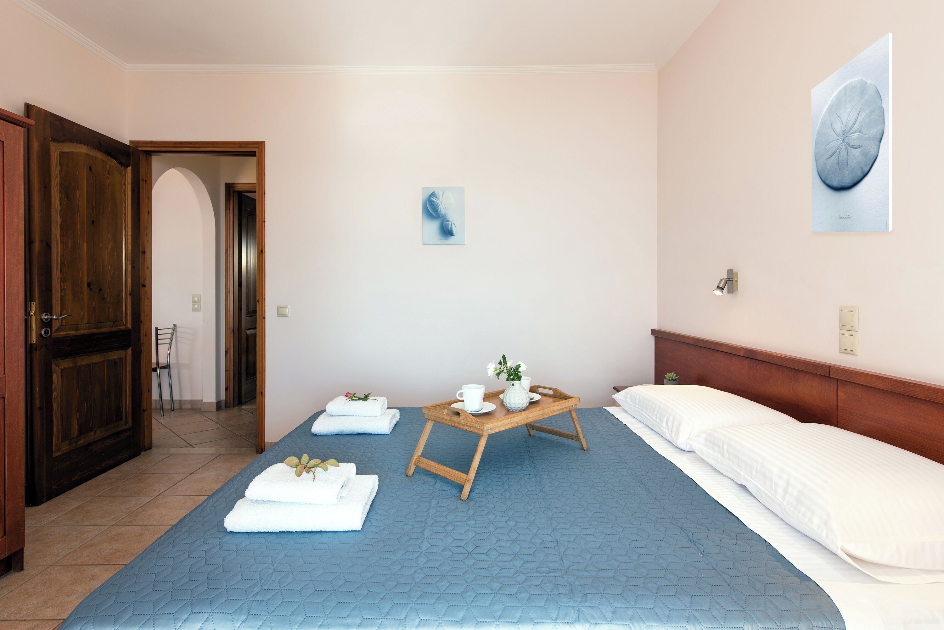 Quarto de hotel com cama, mesa de cabeceira de madeira, uma xícara e um vaso, além de uma luminária na parede.