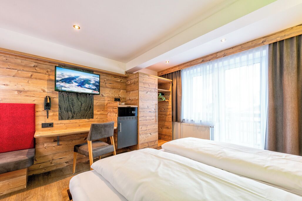 Quarto de hotel com parede de madeira, TV fixada na parede, escrivaninha com cadeira, cama e janela com cortinas.