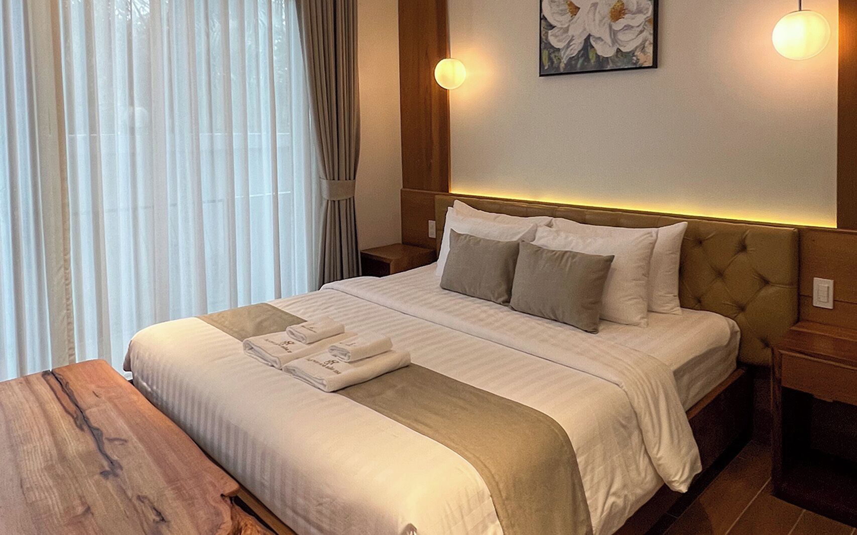 Quarto de hotel com uma cama bem arrumada, mesa de cabeceira de madeira e luminária fixada na parede.