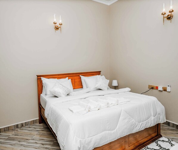 Cama bem arrumada com lençóis brancos e cabeceira de madeira, mesa de cabeceira com abajur, e tomada na parede.