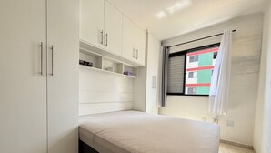 Quarto com cama, guarda-roupa branco e janela com vista para edifícios.
