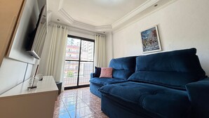Sala de estar com sofá azul, televisão de tela plana na parede, piso de azulejos e janela com cortinas.