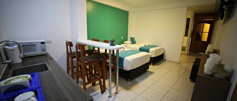 Quarto de hotel compacto com cozinha americana, área de refeições e cama com parede de destaque verde.