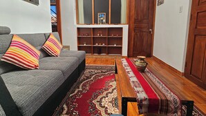 Uma sala de estar com um sofá cinzento, almofadas coloridas, uma mesa de centro de madeira, um tapete estampado em vermelho e uma estante de madeira com objetos decorativos.