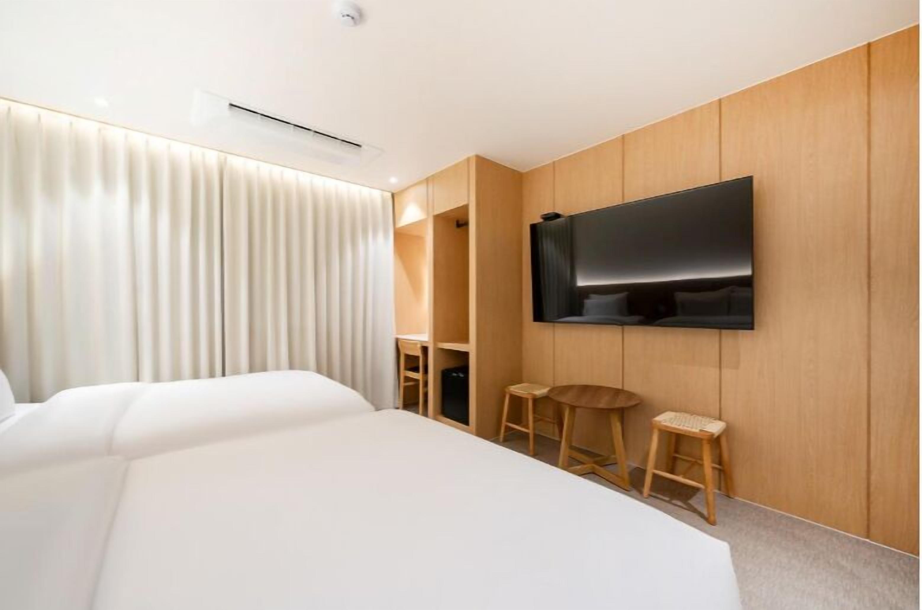 Quarto de hotel moderno com uma cama grande, uma televisão de tela plana na parede e uma pequena área de estar com bancos.