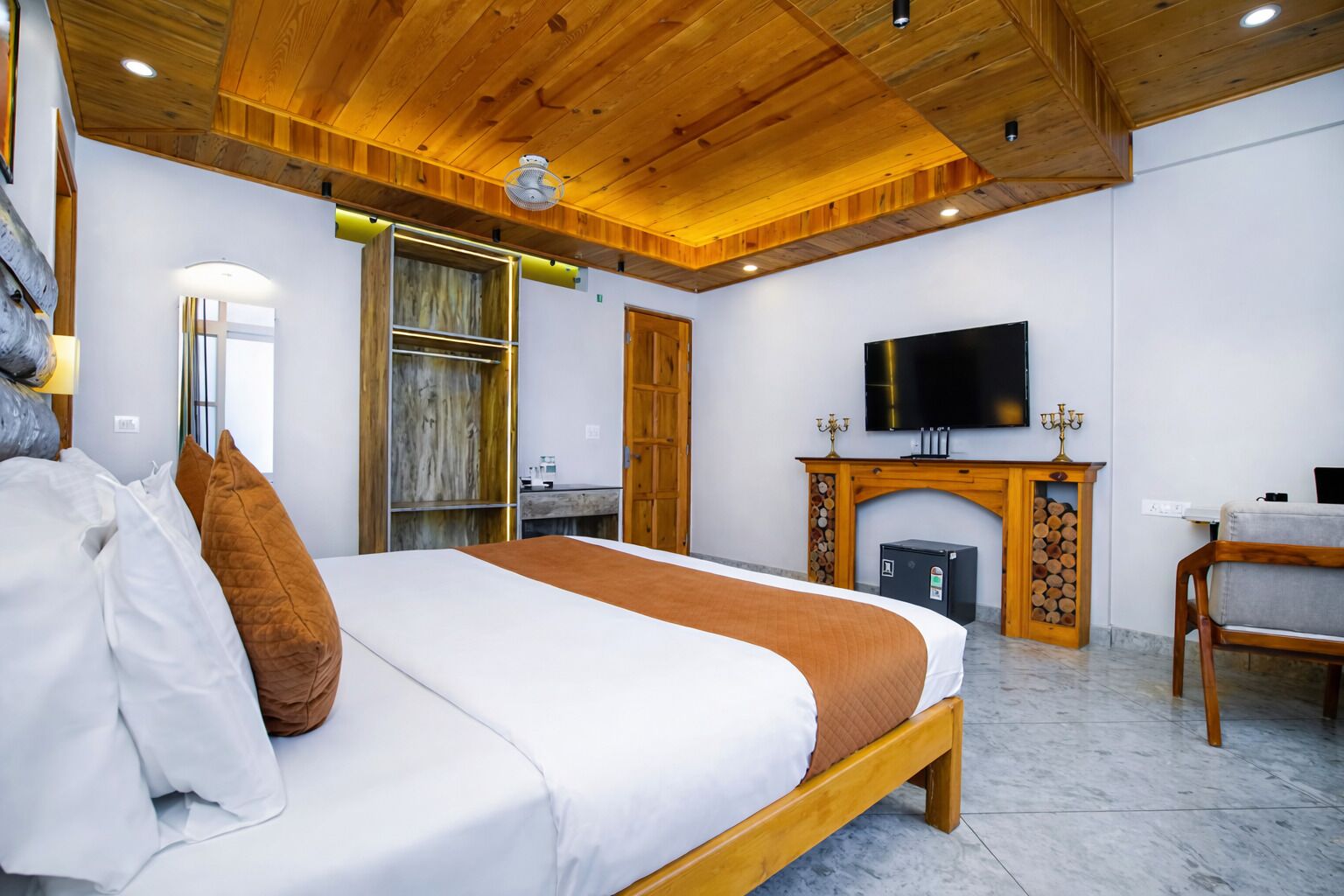 Quarto de hotel moderno com uma cama grande, televisão, lareira e teto de madeira.