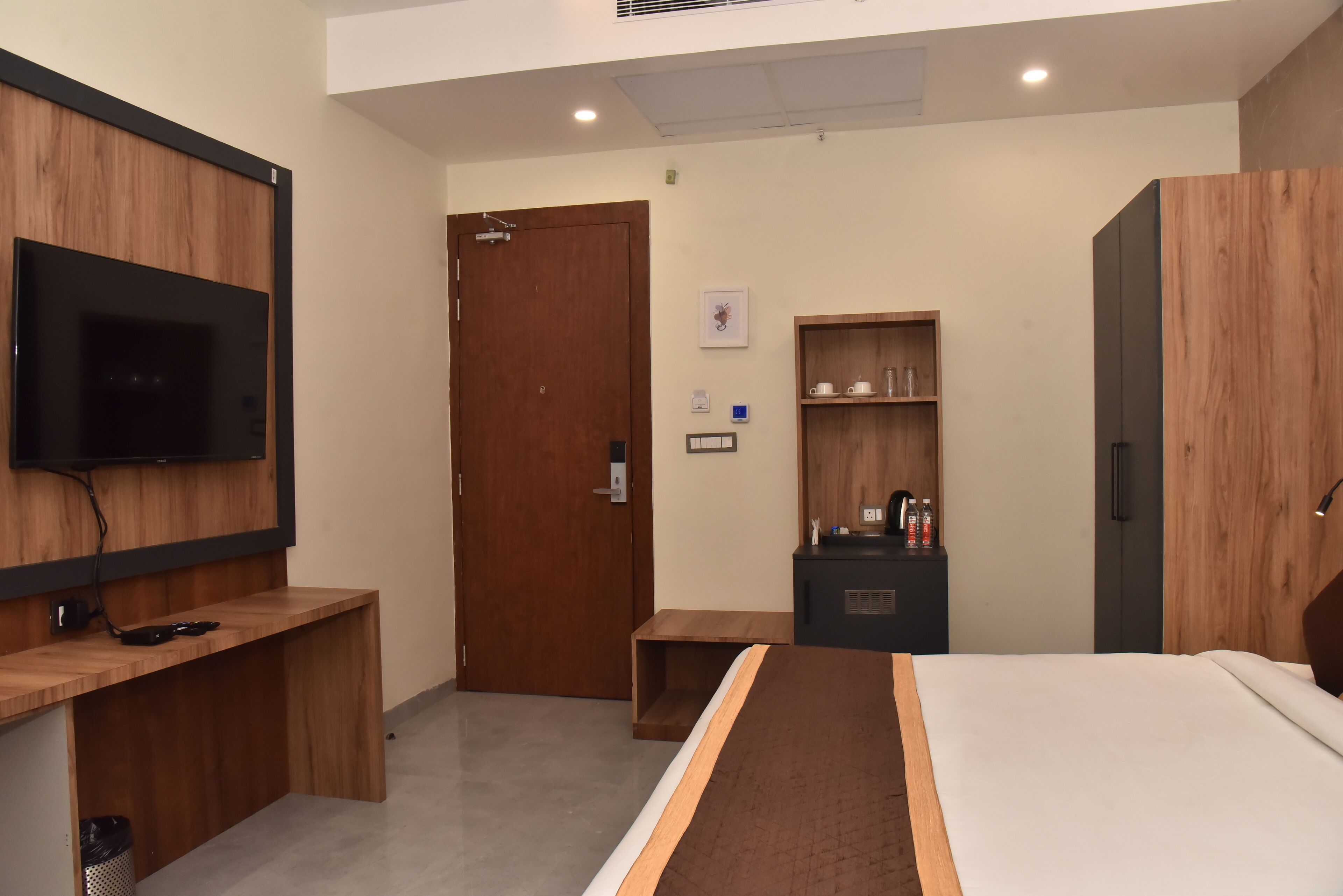 Quarto de hotel com cama, escrivaninha, televisão, armário, prateleira pequena com produtos de higiene e uma porta.