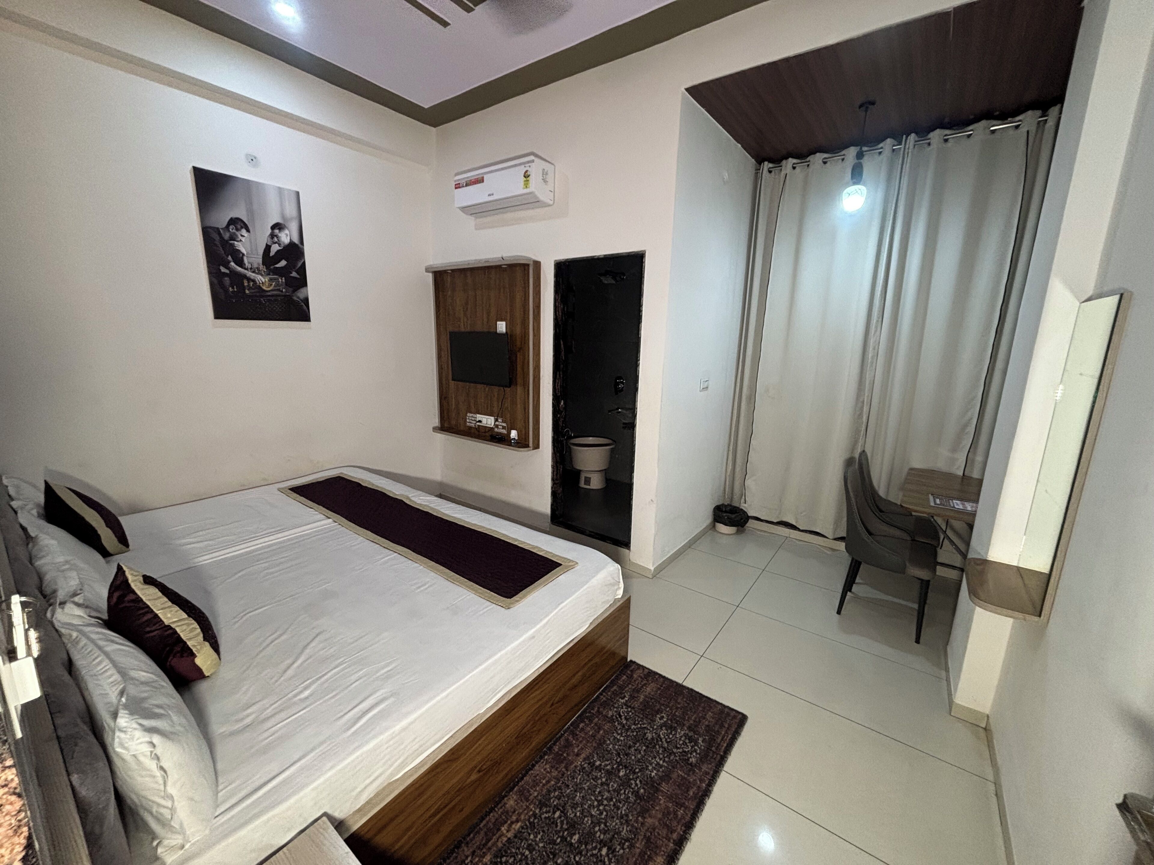 Quarto com cama, escrivaninha, cadeira, televisão fixada na parede e banheiro visível por uma porta aberta.