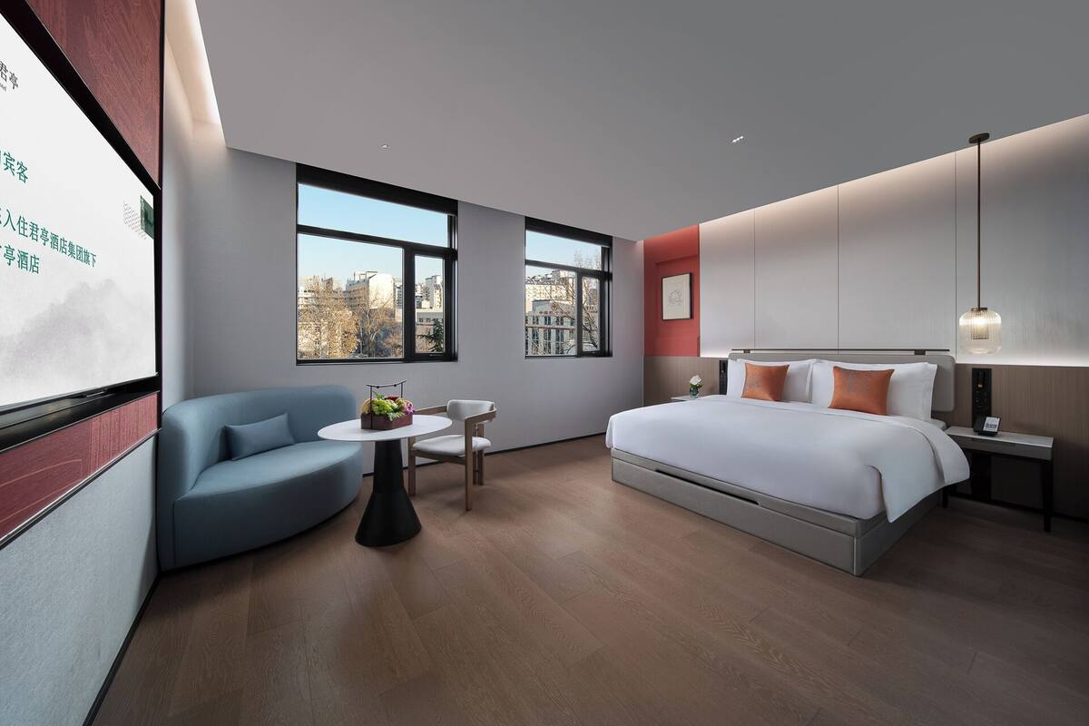 Quarto de hotel moderno com uma cama grande, área de estar com mesa e duas janelas amplas com vista para a cidade.