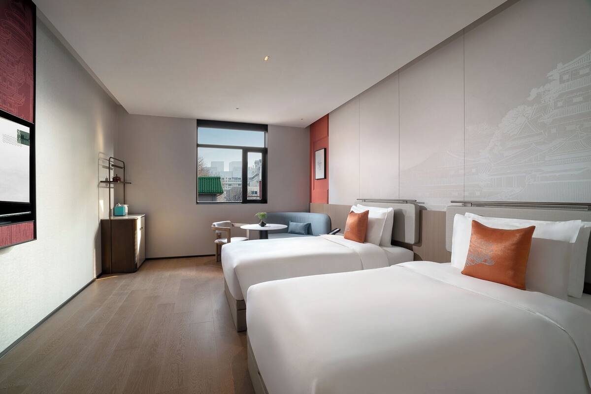Quarto de hotel moderno com duas camas, uma mesinha e uma janela com vista para a cidade.