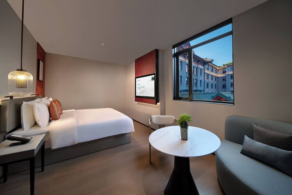 Quarto de hotel moderno com cama, escrivaninha, sofá e janela com vista para arquitetura tradicional.