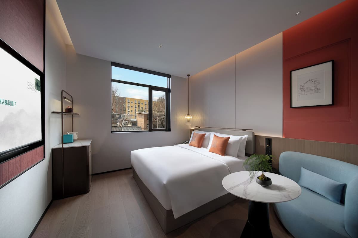 Quarto de hotel moderno com uma cama grande, uma mesinha com uma planta, uma poltrona azul e uma janela com vista para a cidade.