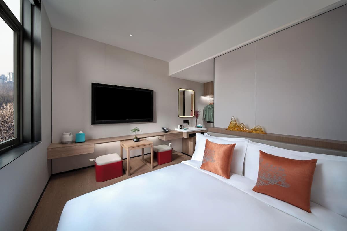 Quarto de hotel moderno com uma cama grande, uma escrivaninha, uma TV de tela plana, um espelho e uma janela com vista para a cidade.