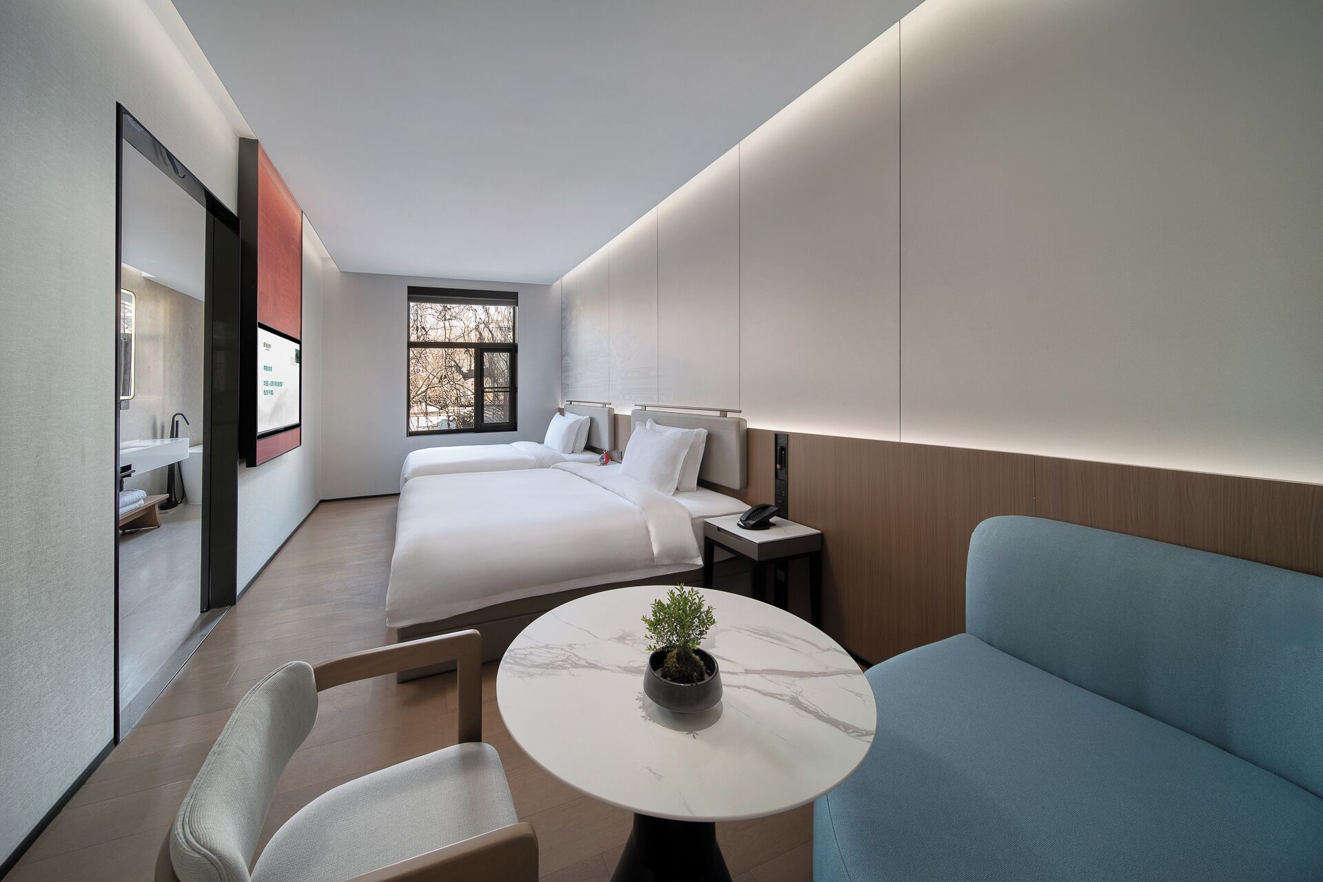Quarto de hotel moderno com duas camas, um sofá, uma pequena mesa com uma planta e um banheiro com pia e espelho.