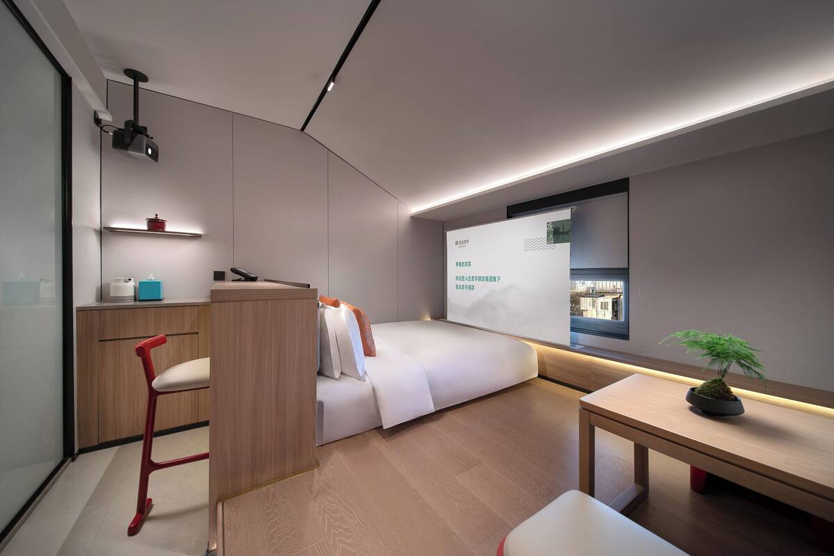 Quarto de hotel moderno e compacto com uma cama, escrivaninha, cadeira, uma mesinha com uma planta e uma janela grande com vista para a cidade.