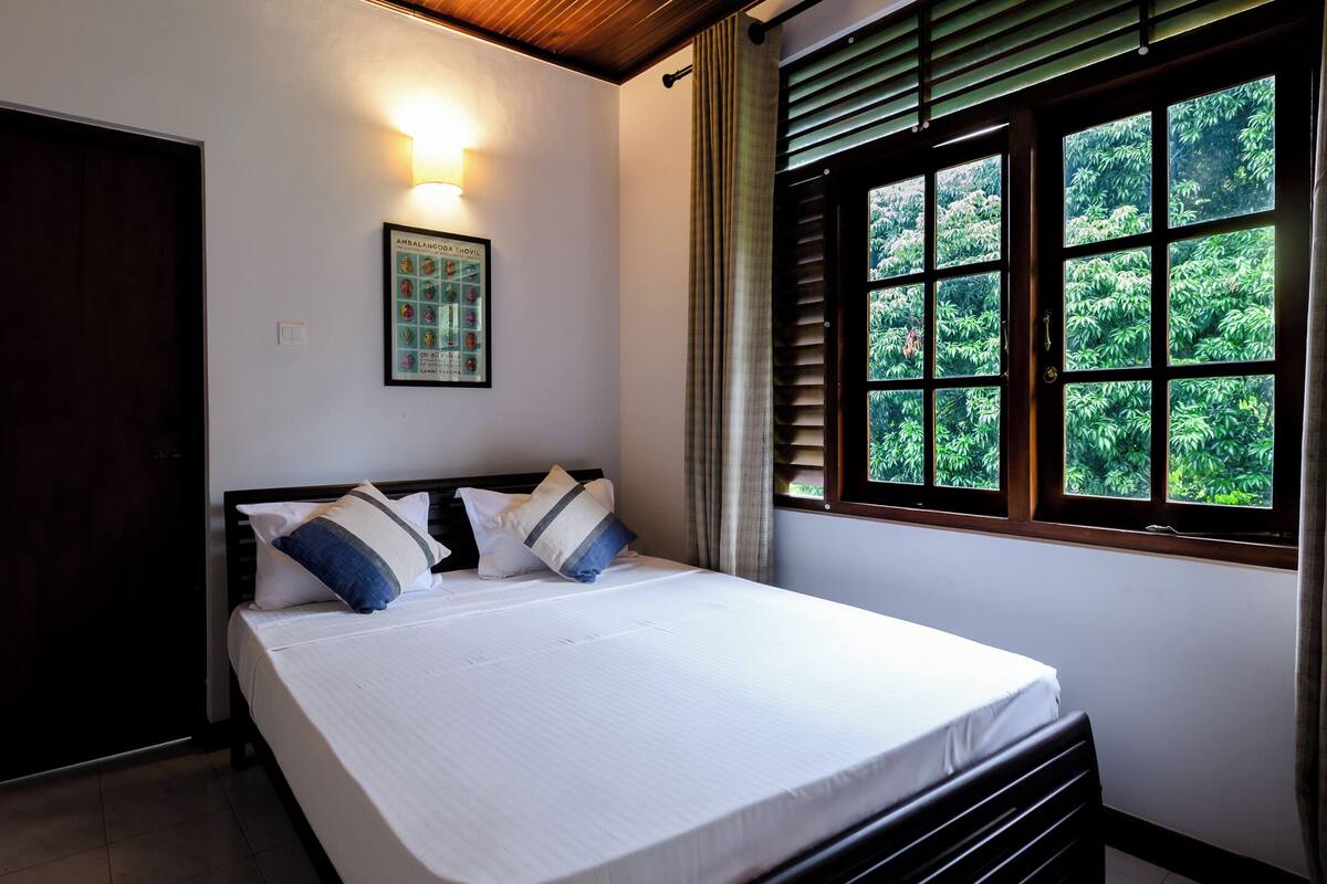 Cama bem arrumada com lençóis brancos e várias almofadas, um quadro na parede e uma janela com vista para o verde.