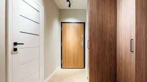 Um corredor com uma porta branca à esquerda, uma porta de madeira no centro e outra porta de madeira à direita.