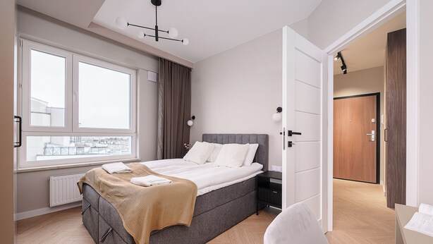 Quarto moderno com uma cama grande, mesa de cabeceira, uma cadeira e vista para a cidade pela janela.