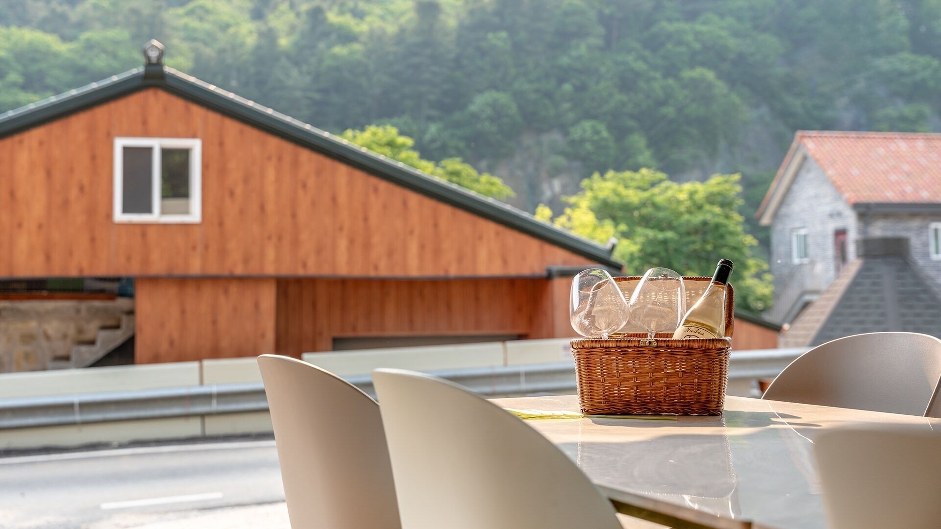 Uma mesa com uma cesta de vime contendo copos de vinho e uma garrafa, com um edifício de madeira e uma parede de pedra ao fundo.