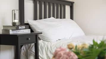 Uma cama bem arrumada com roupa de cama branca, uma mesinha de cabeceira com abajur e alguns livros.